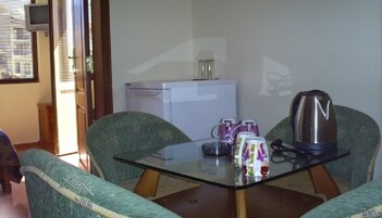 Uma pequena área de refeições com mesa de vidro, duas cadeiras estofadas verdes, um frigorífico branco e uma chaleira sobre a mesa.