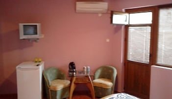 Um quarto pequeno com paredes cor-de-rosa, uma geladeira branca, duas cadeiras verdes, uma mesa com cafeteira e uma janela com persianas.