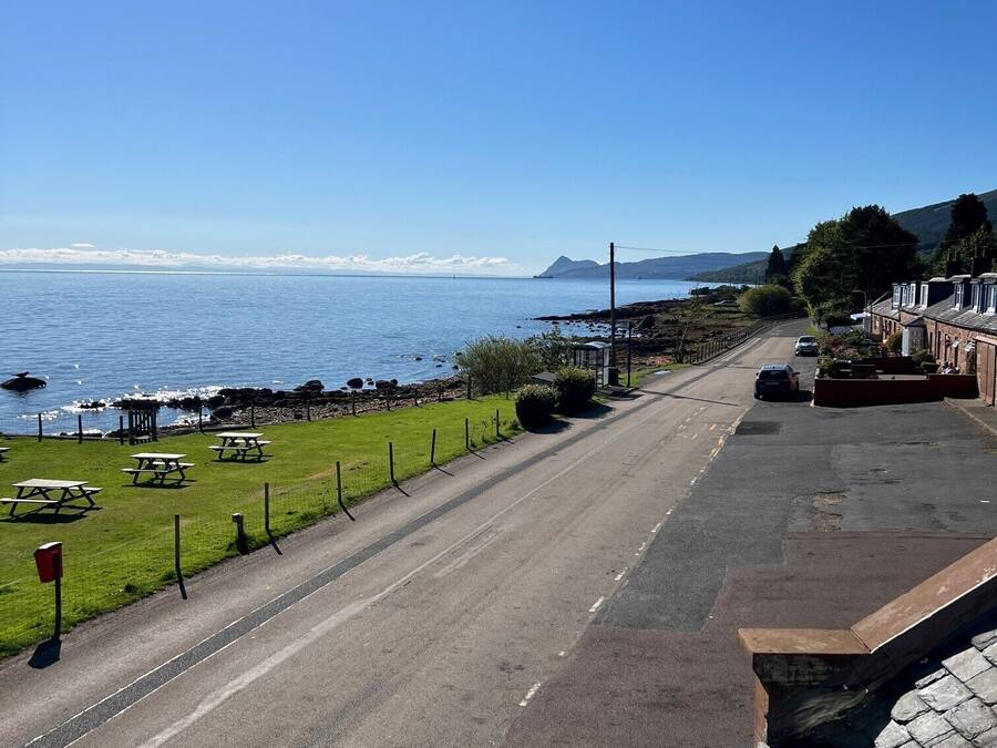 Uma estrada costeira com mesas de piquenique, vista clara do mar e edifícios residenciais.