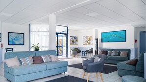 Ampla sala de estar com sofá azul, poltrona cinza e mesa de centro. Há uma janela grande com cortinas leves, um mapa na parede e uma televisão exibindo uma vista panorâmica.