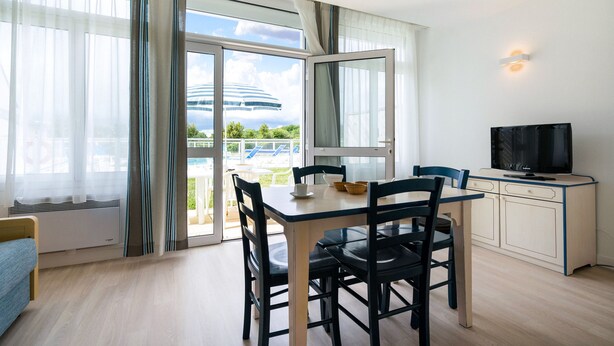 Quarto moderno com mesa de jantar, cadeiras, TV e vista para um edifício por grandes janelas.