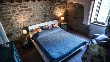 Quarto com parede de pedra, cama com manta azul, mesa de cabeceira com luminária e uma cadeira.