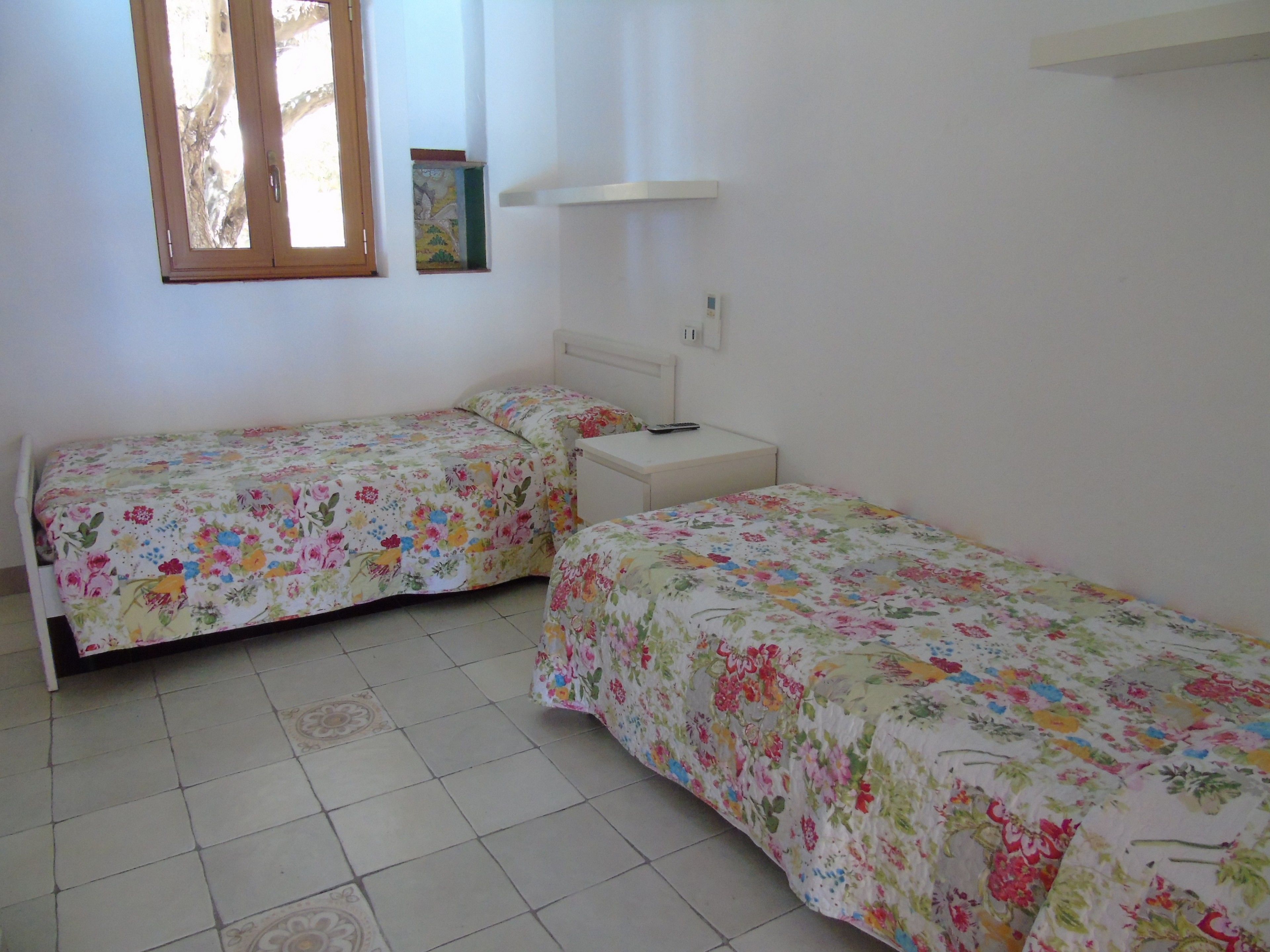 Quarto com duas camas de solteiro, uma pequena mesa de cabeceira com abajur, uma janela e um prateleira na parede.