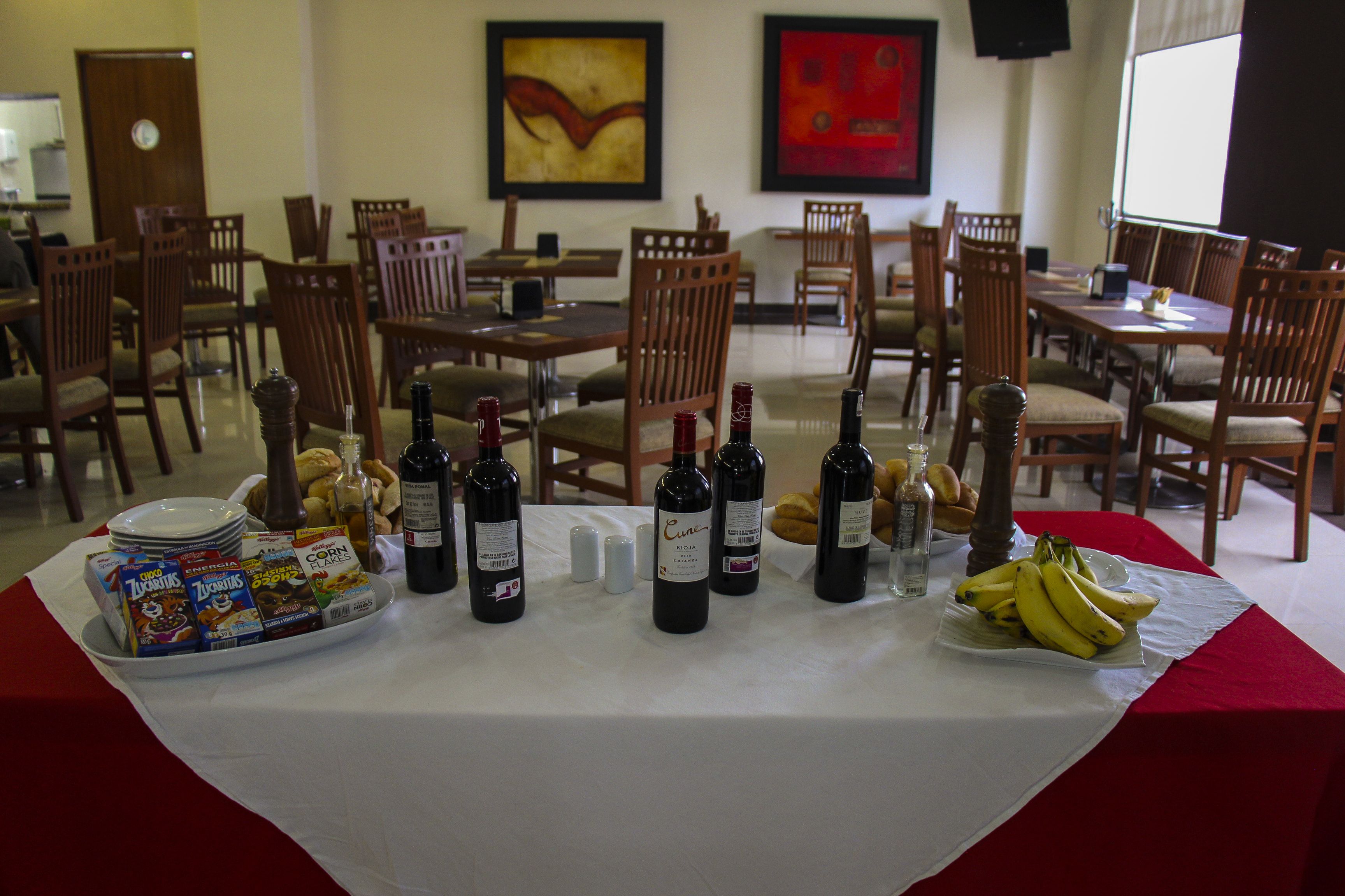Uma sala de jantar com mesas preparadas para uma refeição, com garrafas de vinho, pão e um prato de frutas.