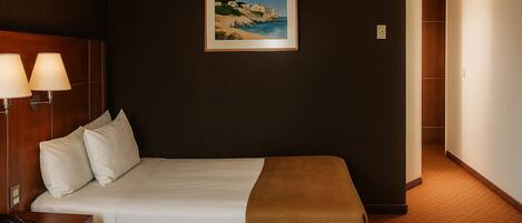Quarto de hotel com cama, abajures, telefone, quadro na parede e corredor que leva a um armário.