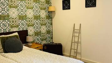 Um quarto arrumado com um edredom estampado, uma luminária de cabeceira, uma mala e uma escada encostada na parede.