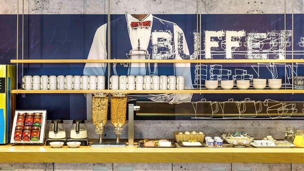 Um balcão de buffet com uma grande placa indicando 'BUFFET' e um uniforme de chef. Há várias estações de serviço com comida e bebidas.