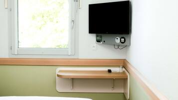 Um quarto de hospital com uma cama, uma janela com vista para o verde, uma televisão montada e uma pequena prateleira.