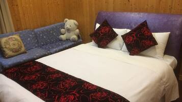 Uma cama com roupa de cama branca e um cobertor estampado com rosas pretas e vermelhas. Há um sofá azul com um ursinho de pelúcia e uma almofada decorativa.