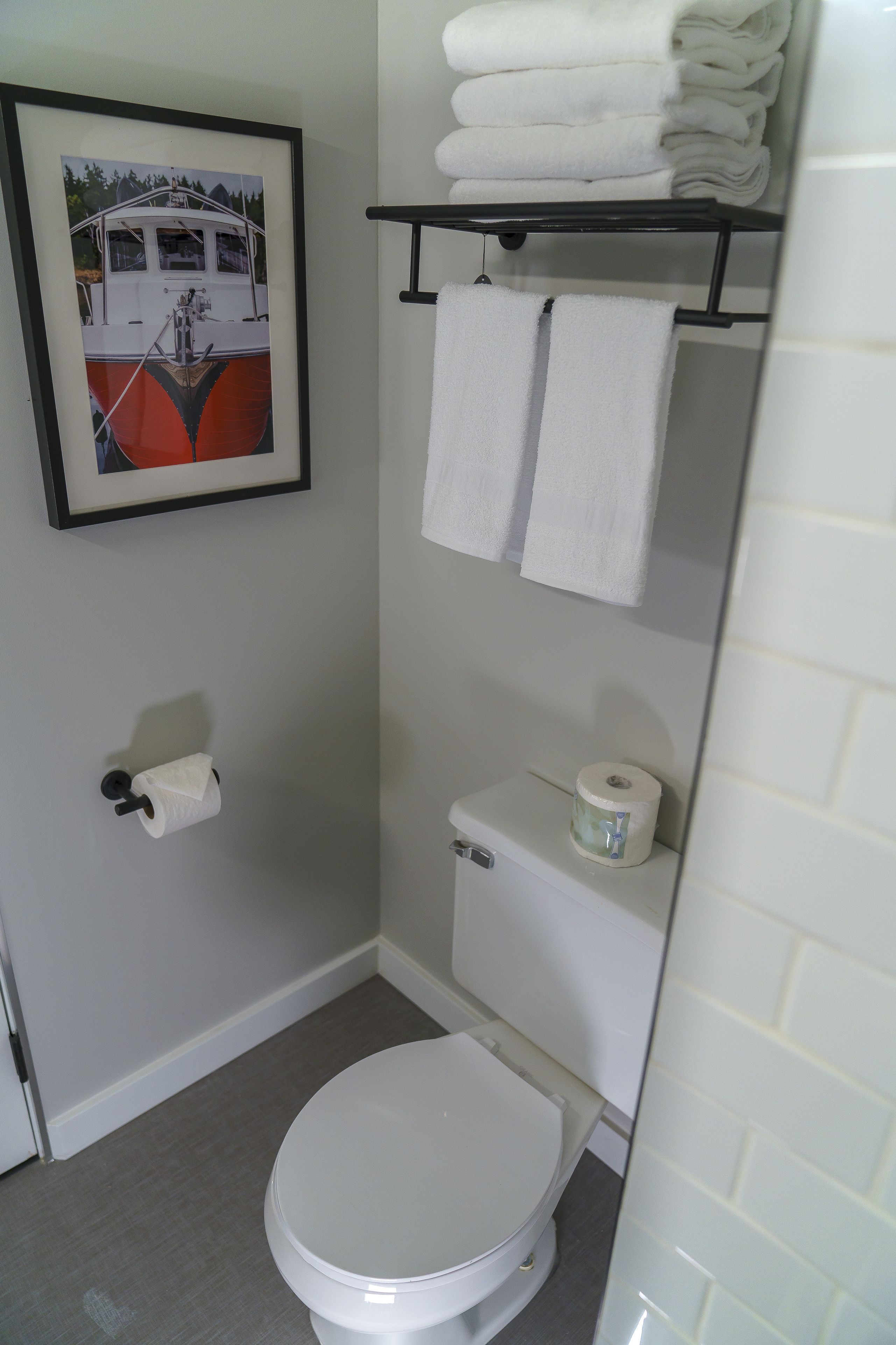 Casa de banho com sanita, um quadro na parede, uma pilha de toalhas brancas e rolo de papel higiénico.