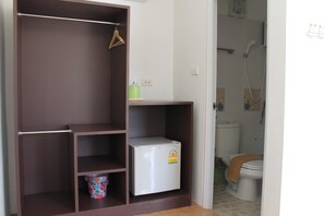 Quarto compacto com guarda-roupa, pequeno frigorífico e casa de banho visível através de uma porta aberta.