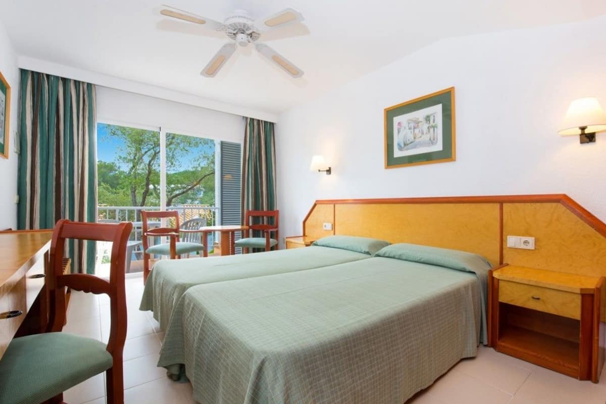 Quarto de hotel com duas camas, uma escrivaninha, cadeiras, ventoinha no teto e uma janela com cortinas.