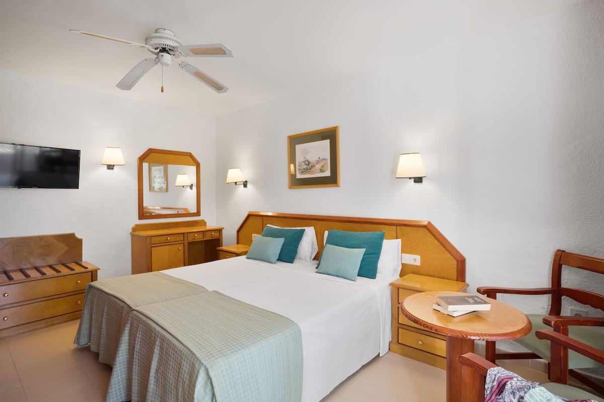 Quarto de hotel com cama, escrivaninha, cadeira, ventilador de teto, televisão e luminárias fixadas na parede.