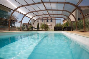 Piscina transparente com estrutura de vidro, rodeada por plantas e um edifício ao fundo.