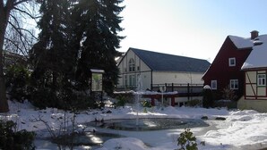 Área externa coberta de neve com um lago, uma grande árvore perene e edifícios com arquitetura tradicional.