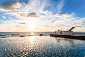 Uma piscina infinita com vista para o pôr do sol, cadeiras de lounge e um deck de madeira.