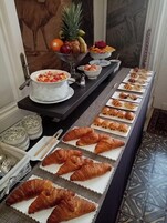 Mesa de buffet com diversos alimentos, incluindo frutas, croissants e uma tigela de legumes variados.