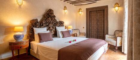 Quarto com uma cama grande, cabeceira de madeira, arandelas decorativas, uma porta de madeira e uma cadeira.