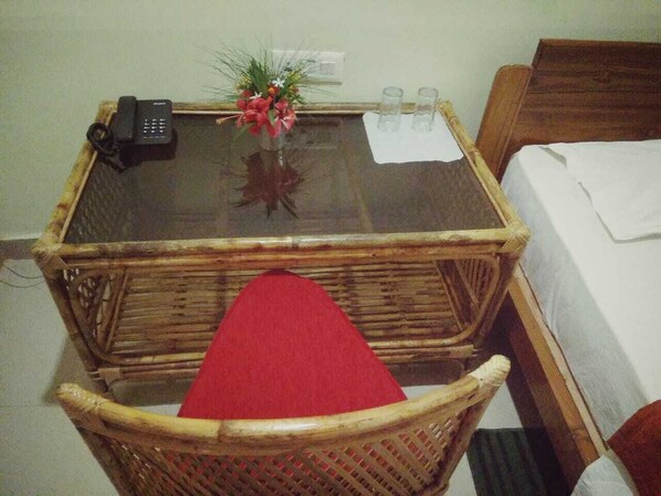 Um quarto pequeno com uma mesa de vidro, uma cadeira de vime, uma cama, uma mesinha de cabeceira, um telefone e um vaso com flores.