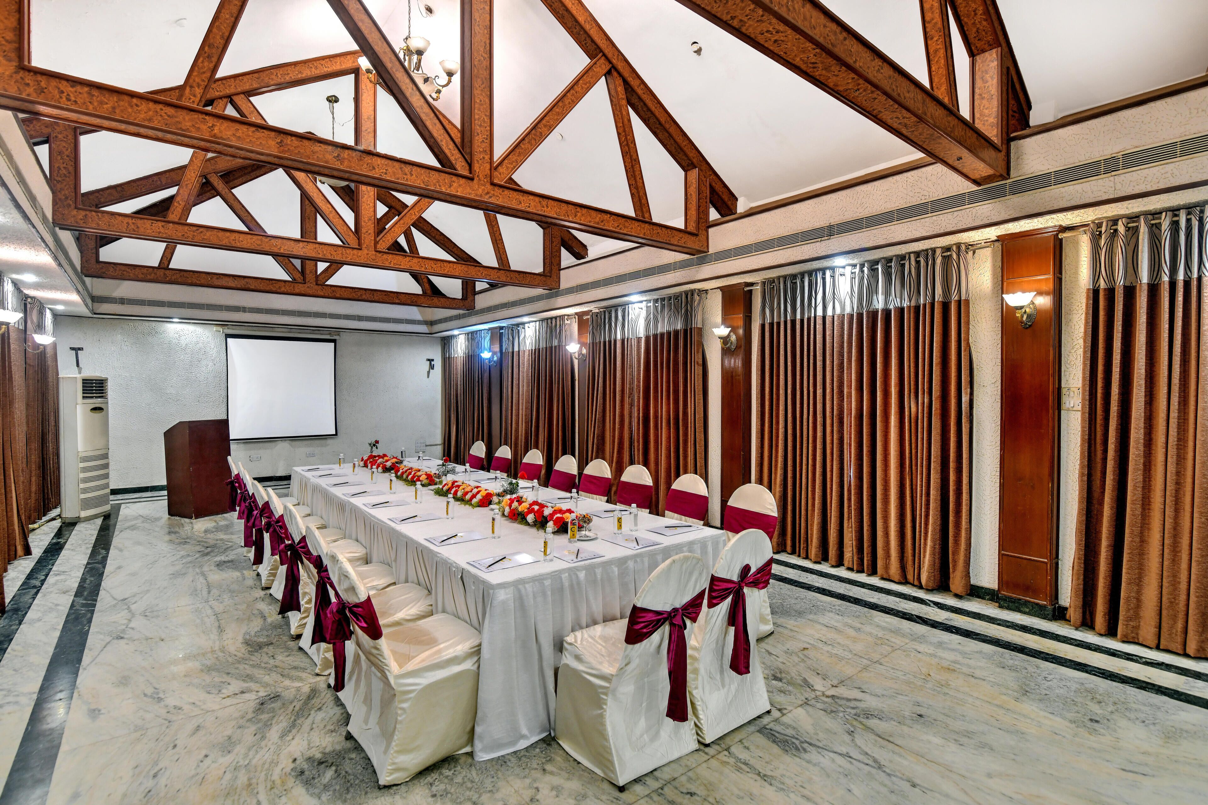 Um salão de banquetes com uma longa mesa posta para um evento formal, vigas de madeira no teto e cortinas nas paredes.