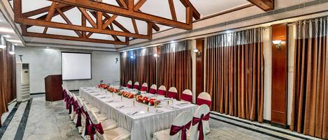 Um salão de banquetes com uma longa mesa posta para um evento formal, vigas de madeira no teto e cortinas nas paredes.