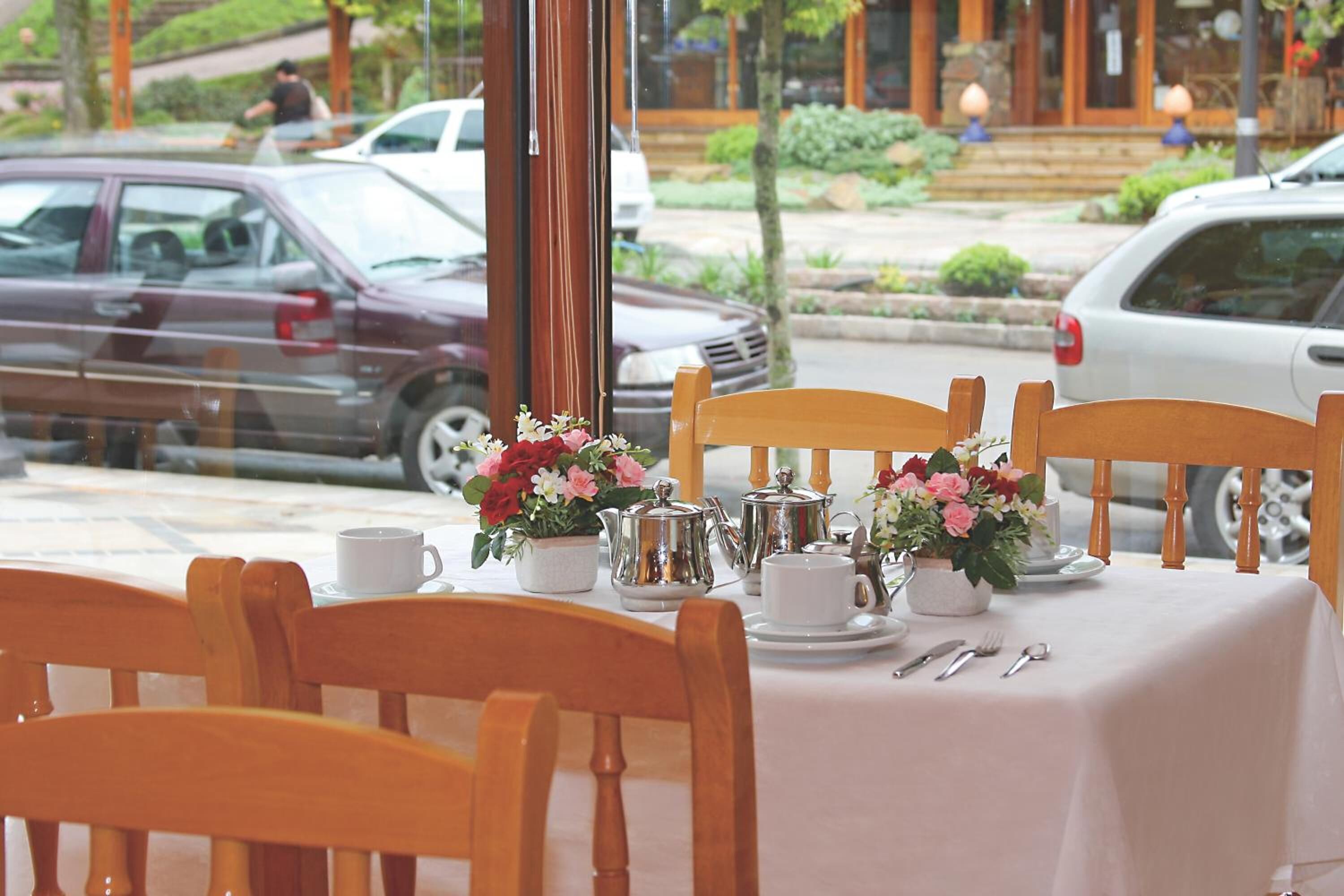 Mesa posta para refeição, com cadeiras, toalha de mesa e vista para carros estacionados e vegetação lá fora.
