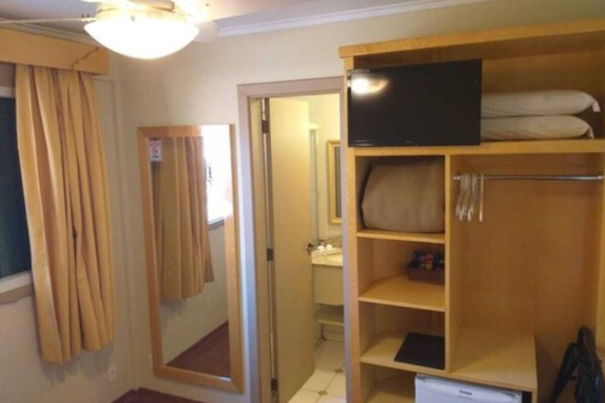 Quarto compacto com guarda-roupa de madeira, televisão por cima e um pequeno frigorífico.