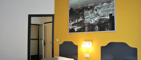 Quarto de hotel com duas camas, uma mesinha de cabeceira com telefone, uma luminária fixada na parede e uma grande fotografia da paisagem urbana.