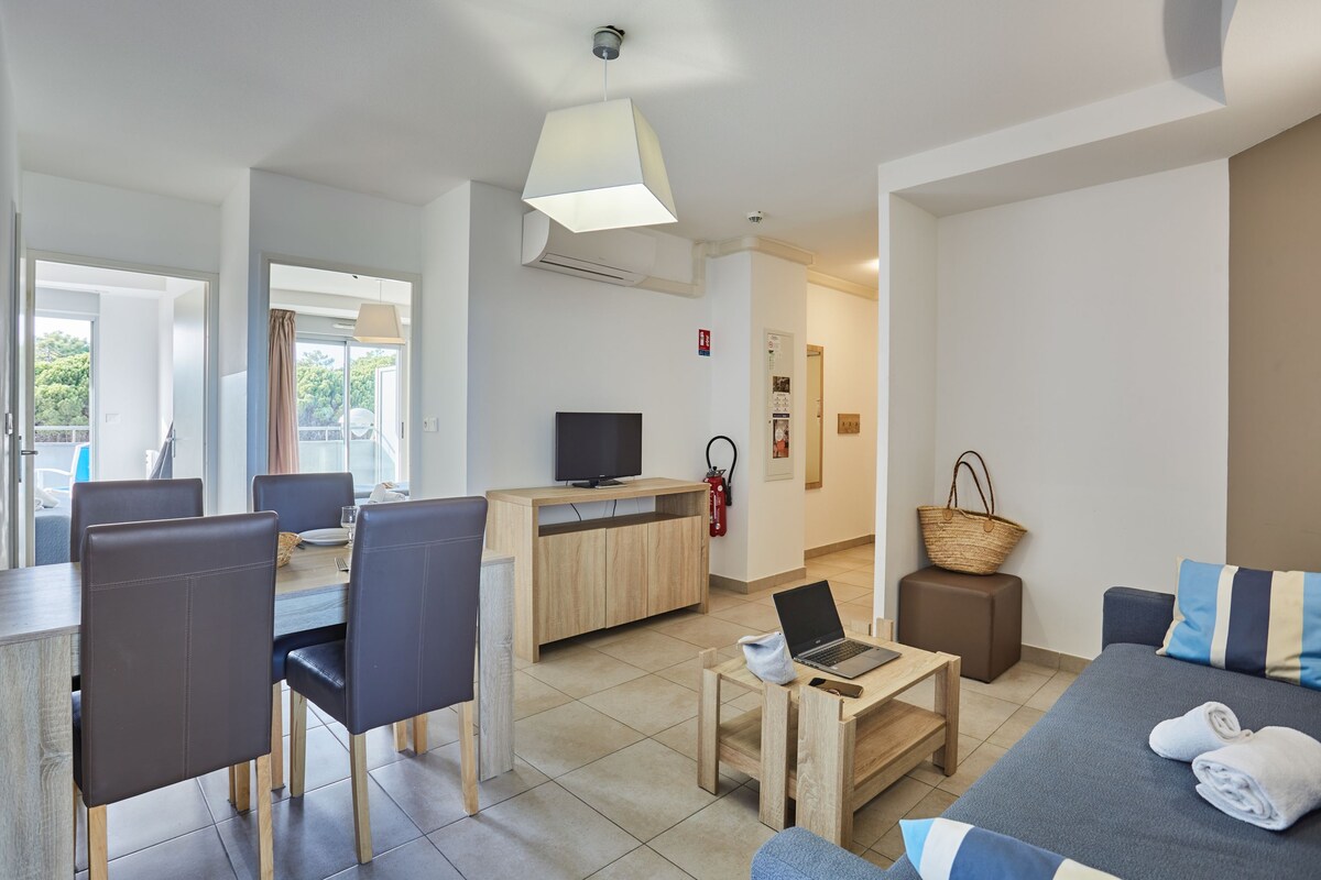 Quarto de hotel moderno com área de jantar, televisão, laptop sobre uma pequena mesa, sofá e toalhas, e extintor de incêndio.