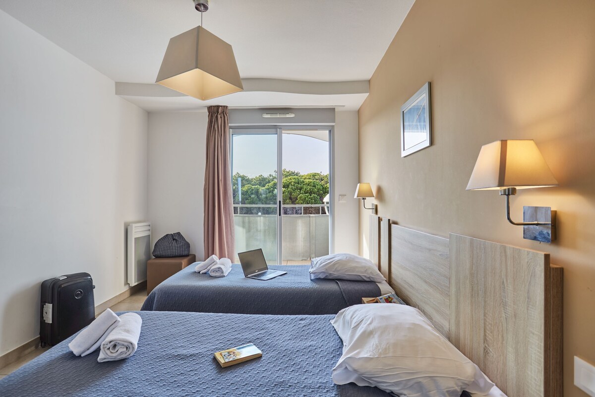 Quarto de hotel com duas camas, um laptop, uma mala e vista para o verde pela janela.