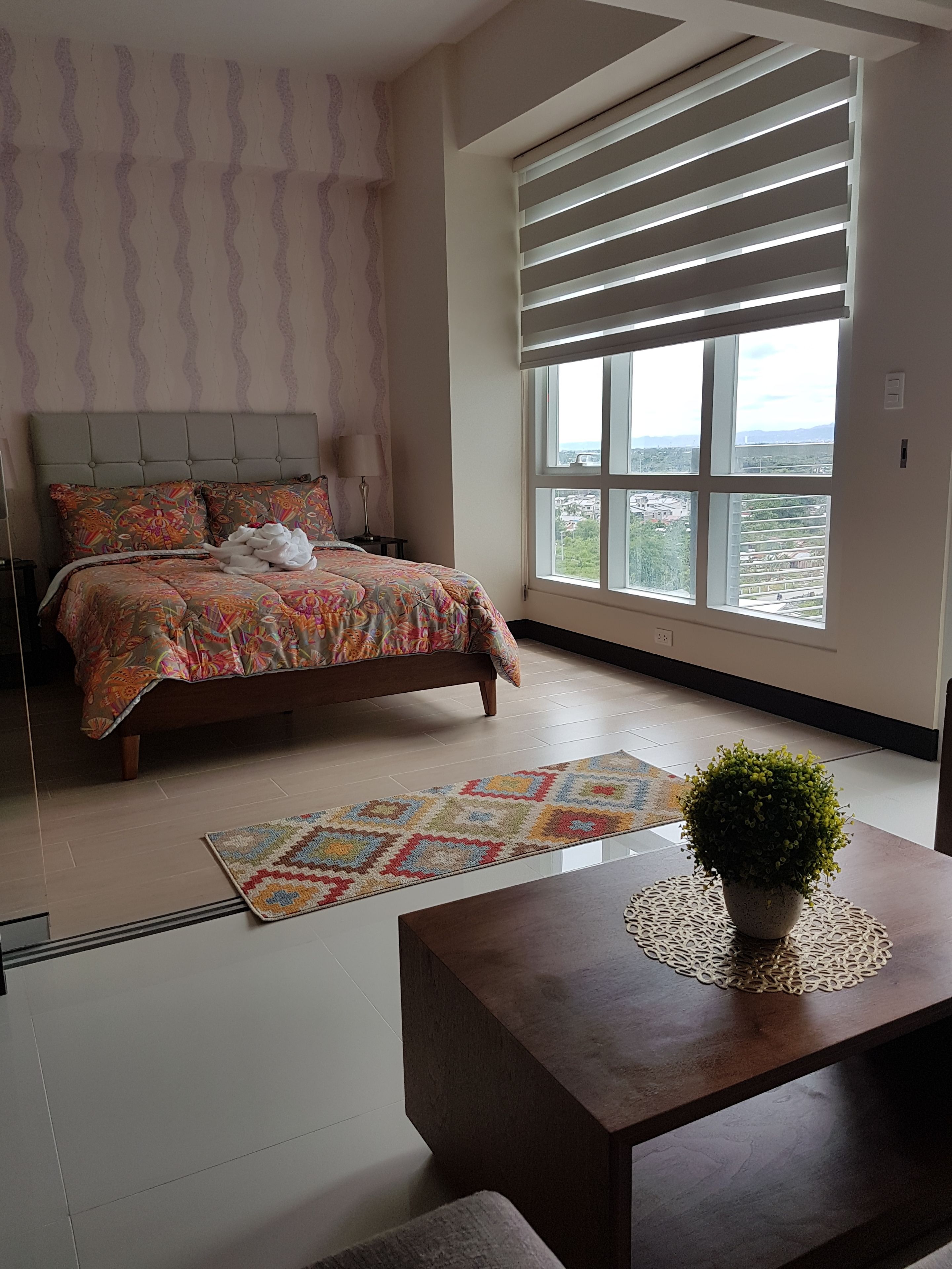 Quarto com cama, mesa de cabeceira, cortinas na janela, planta em vaso e tapete estampado.