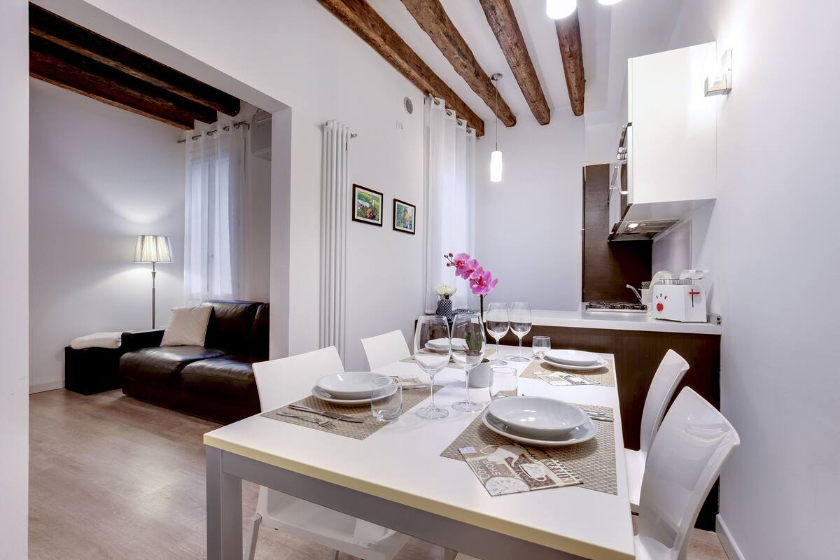 Uma sala de jantar com mesa posta para quatro pessoas, um sofá e uma cozinha ao fundo.