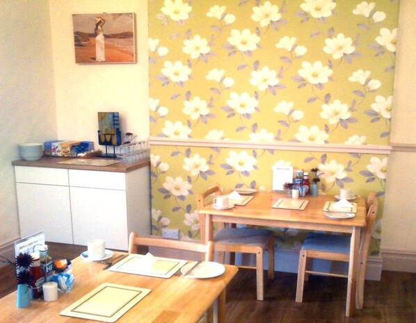 Uma sala de jantar com uma mesa de madeira posta para quatro pessoas, papel de parede floral e um quadro na parede.