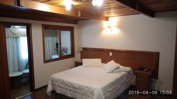 Quarto com cabeceira de madeira, cama com roupa de cama branca, mesa de cabeceira e apoio para luminária. Há um espelho na parede, uma janela com cortinas, e uma pequena mesa com cadeiras fora do quarto.