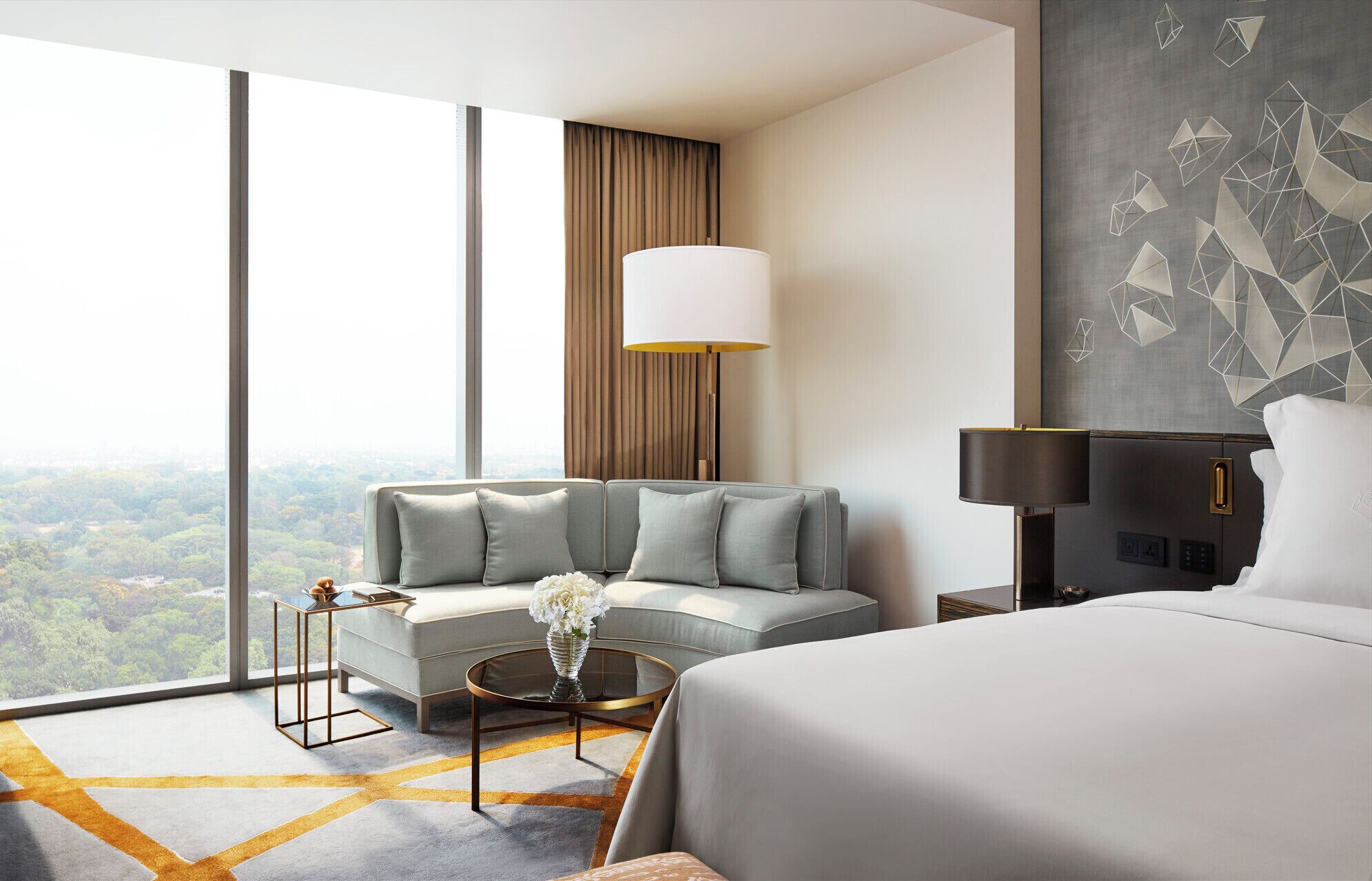 Quarto de hotel moderno com uma grande janela, um sofá, uma cama, uma mesinha de cabeceira e um abajur.