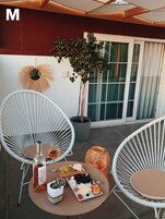 Uma área de pátio com duas cadeiras de vime branco, uma mesa redonda com uma garrafa de vinho, copos e uma tábua de queijos.