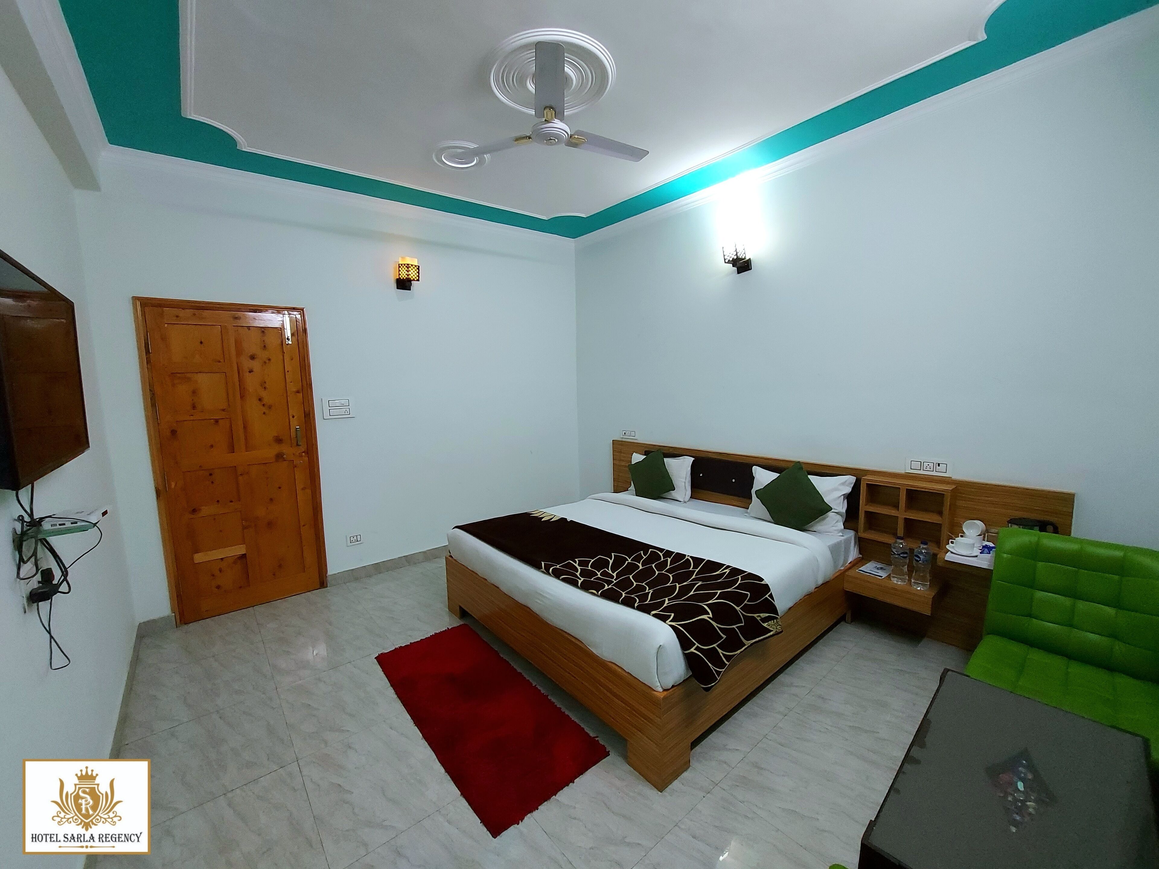 Quarto de hotel com cama, escrivaninha, cadeira verde, tapete vermelho, ventilador de teto e televisão fixada na parede.