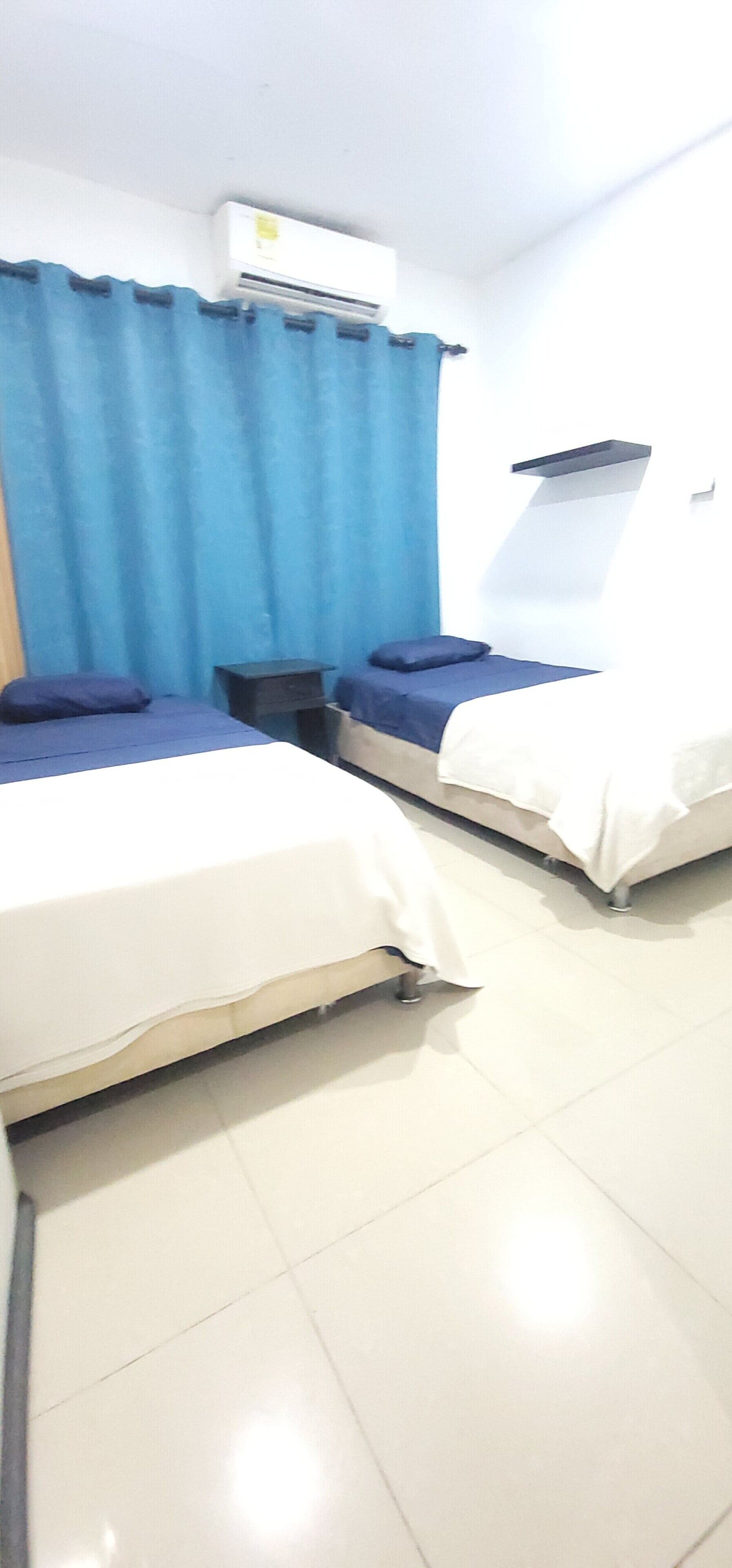 Quarto de hotel com duas camas, uma mesinha, ar-condicionado na parede e uma janela com cortinas.