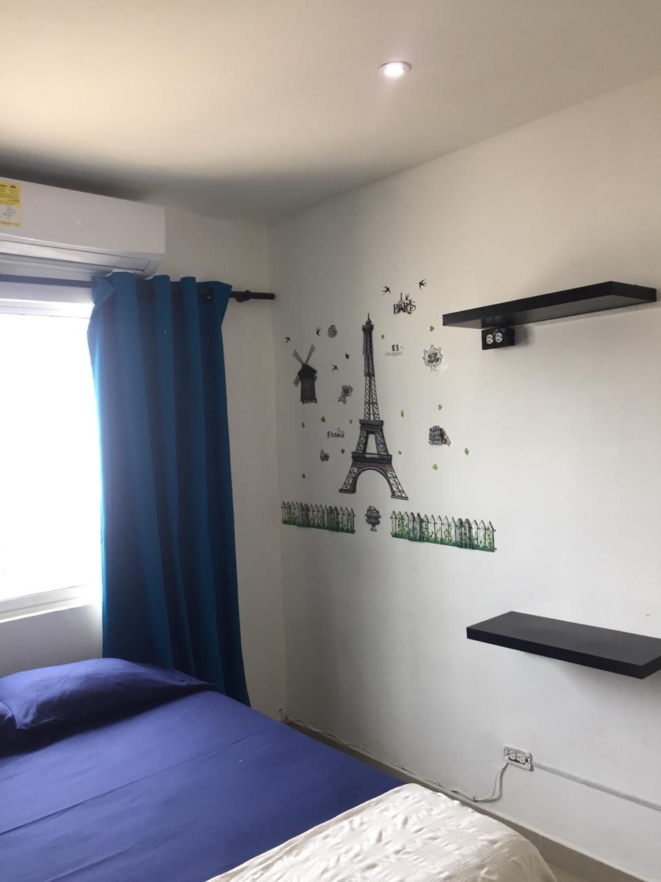 Quarto com cama, janela com cortinas azuis, parede com adesivos da Torre Eiffel e uma prateleira.