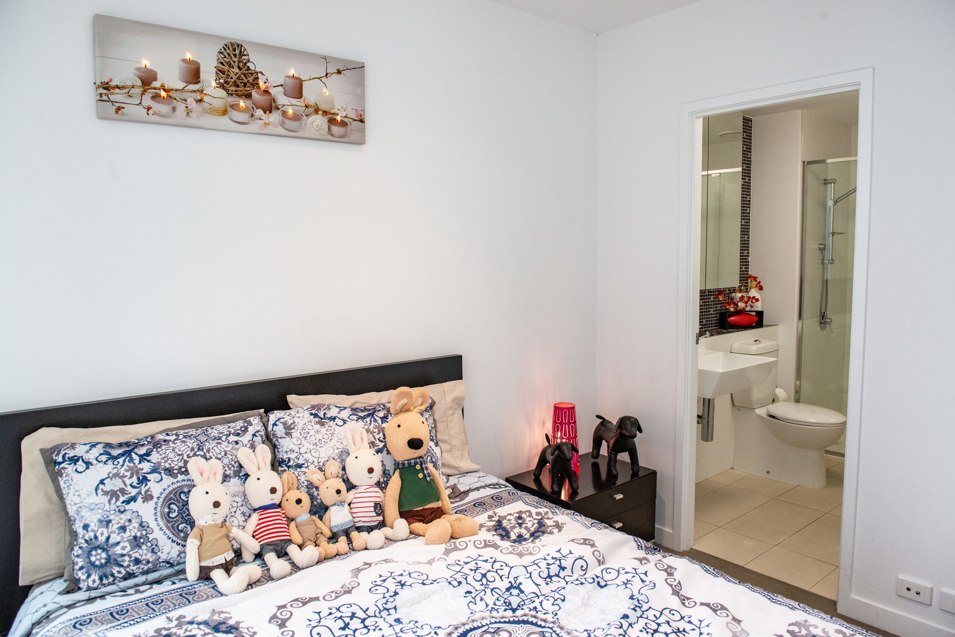 Quarto com cama, brinquedos de pelúcia, decoração na parede e banheiro visível através de uma porta aberta.