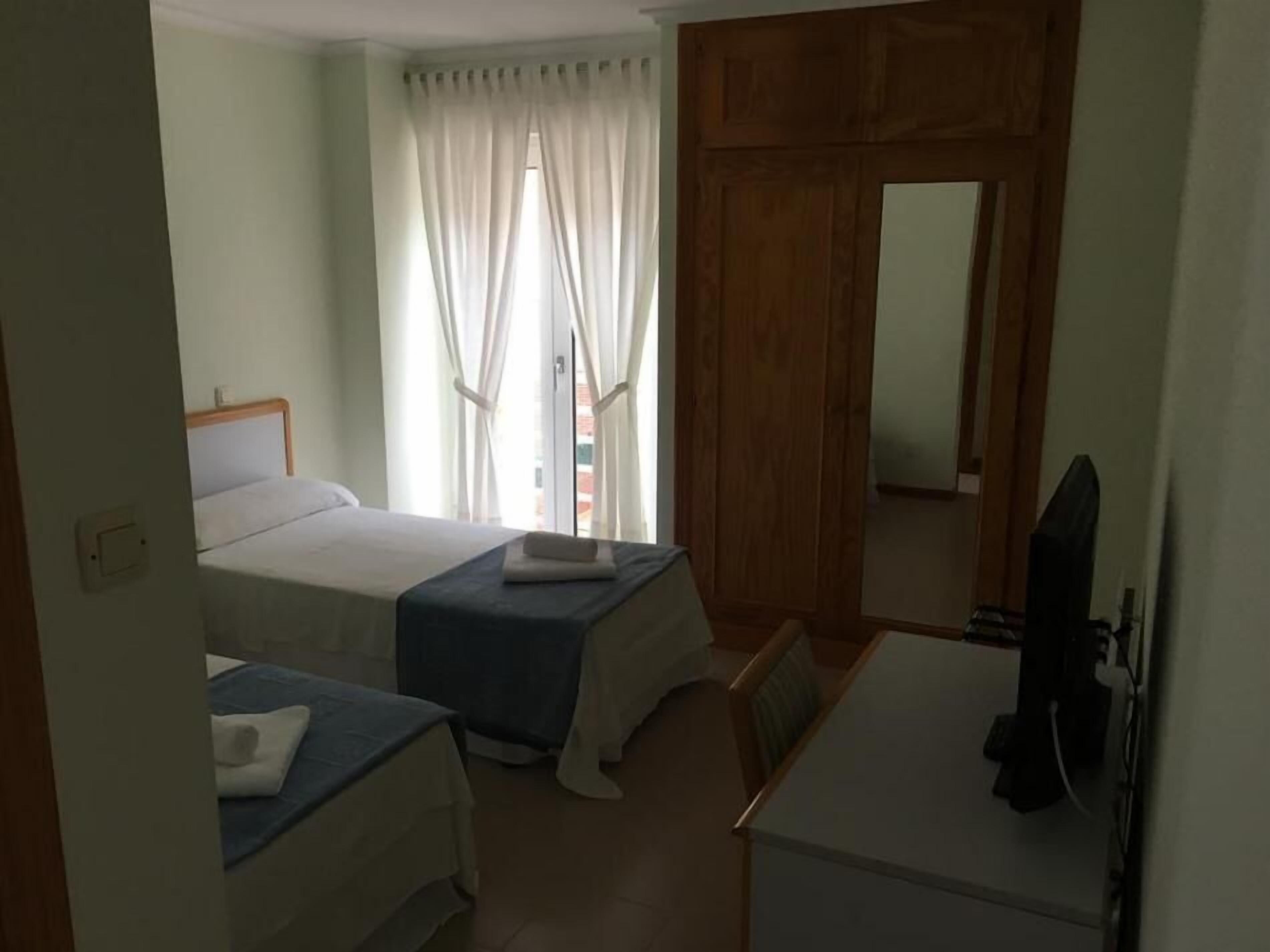 Quarto de hotel com duas camas, uma escrivaninha, uma cadeira, uma televisão e um guarda-roupa.
