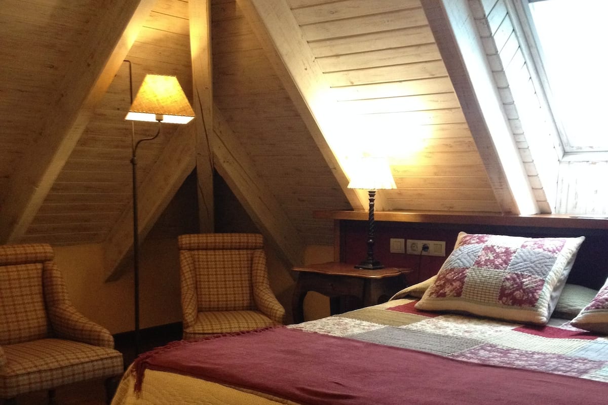 Um quarto de sótão aconchegante, com teto inclinado, uma cama com cobertor vermelho, duas cadeiras xadrez, uma mesa de cabeceira e um abajur.