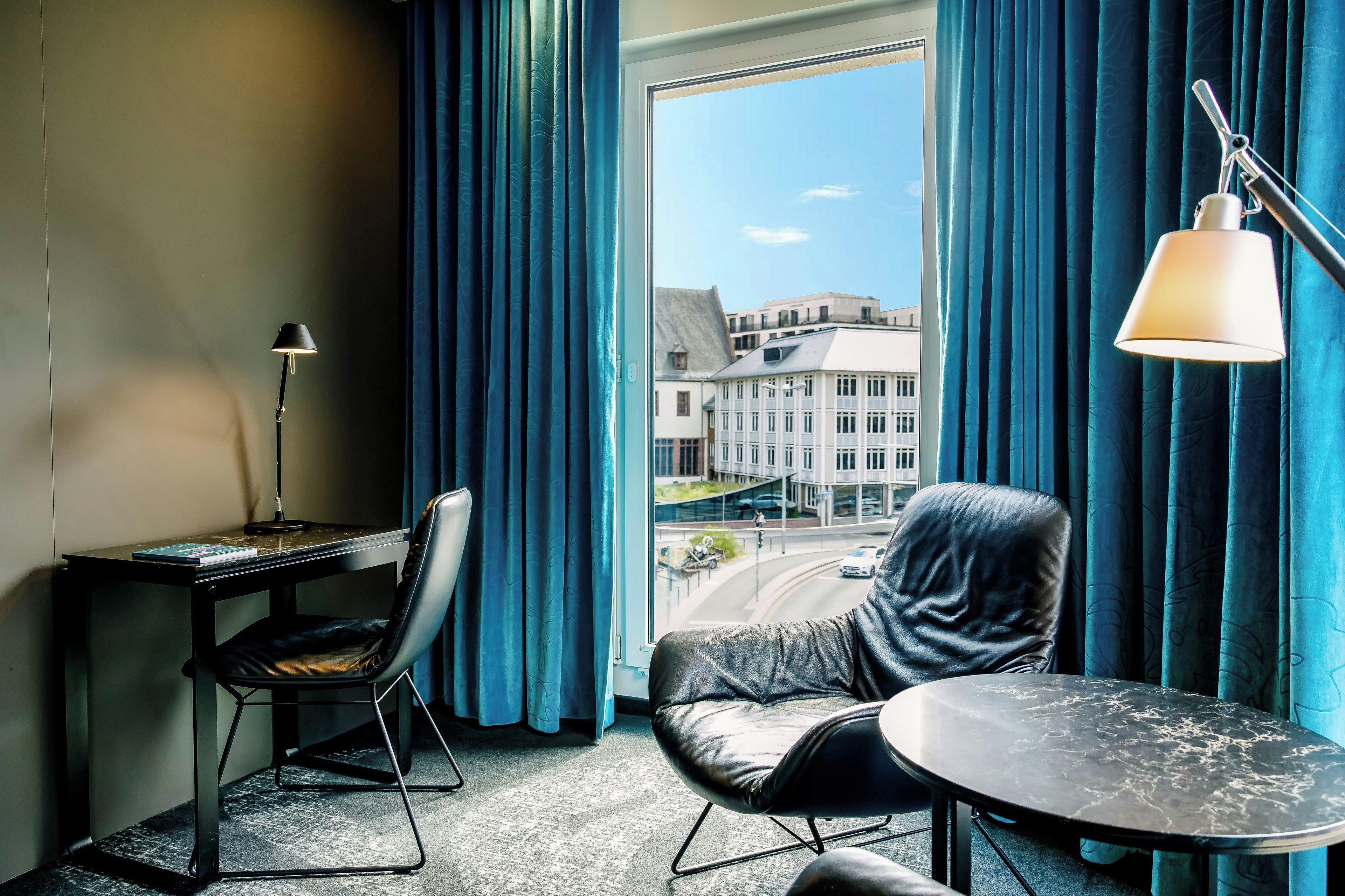 Quarto de hotel com escrivaninha, cadeira e vista para a rua através de uma janela grande.