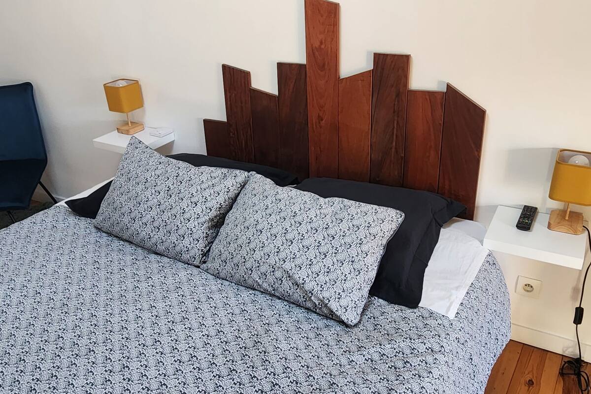Cama bem arrumada com roupa de cama estampada, cabeceira de madeira, mesa de cabeceira com abajur e uma cadeira.