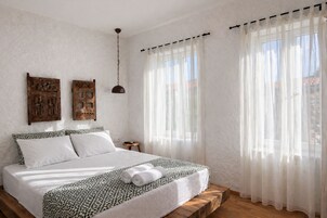 Quarto com cama grande, duas obras de arte na parede, luminária pendente, cortinas translúcidas e um banco de madeira.