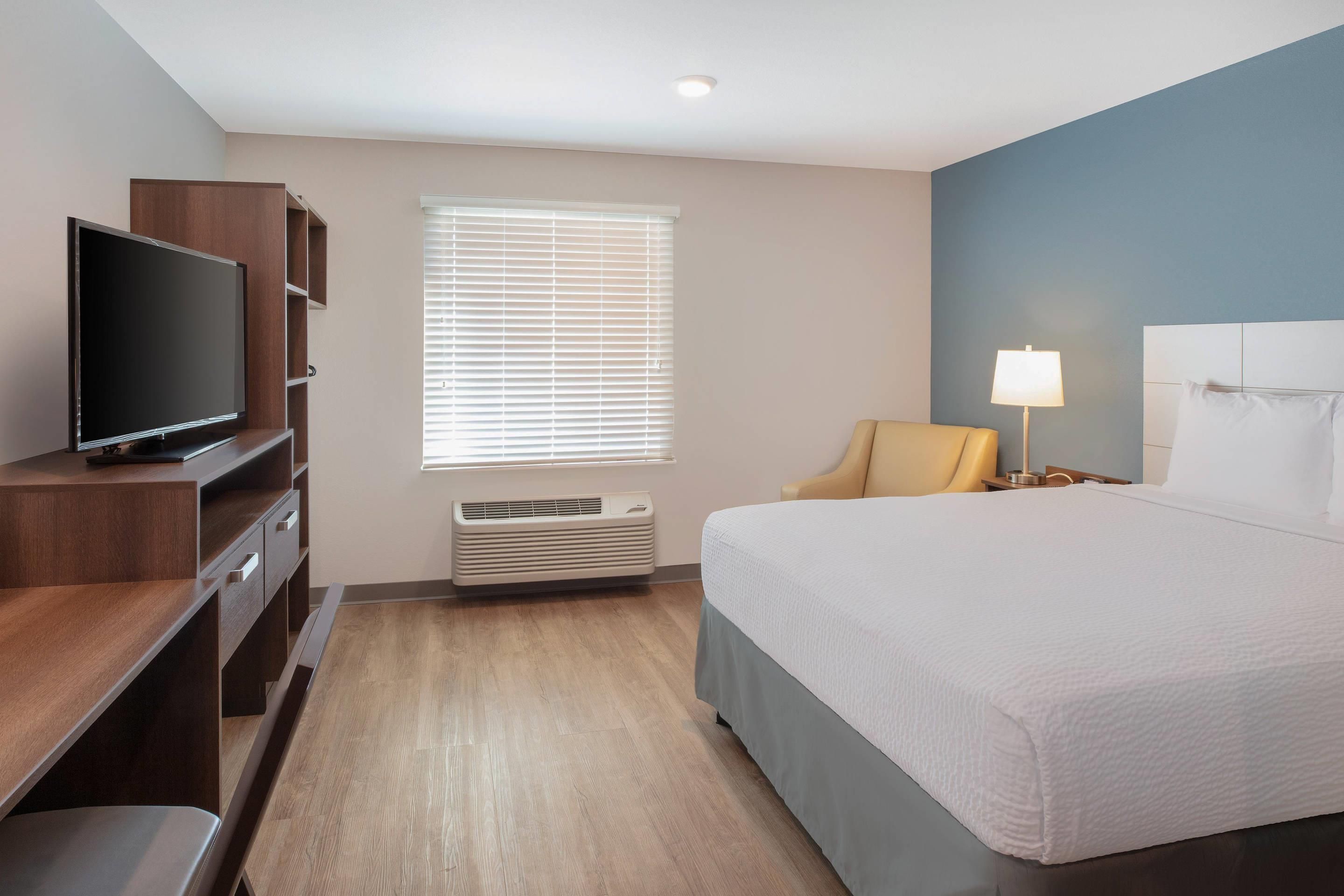 Quarto de hotel com cama, televisão em cómoda de madeira, cadeira, candeeiro e janela com persianas.