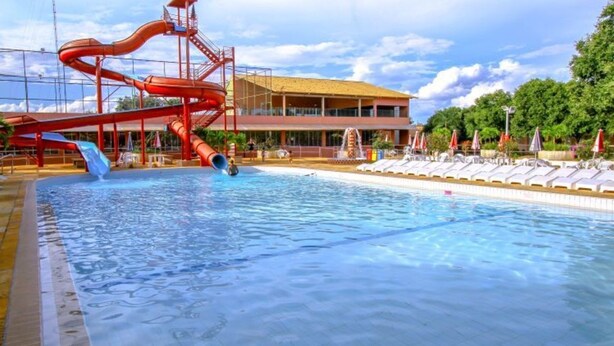 Uma piscina com escorrega, espreguiçadeiras e um edifício com telhado de palha ao fundo.