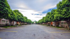 Uma fileira de edifícios baixos com uma área aberta central, possivelmente um estacionamento, cercada por árvores.