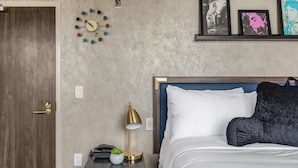 Quarto de hotel com porta de madeira, relógio de parede, mesa de cabeceira com abajur, cama com almofadas brancas e escuras, e quadros na parede.