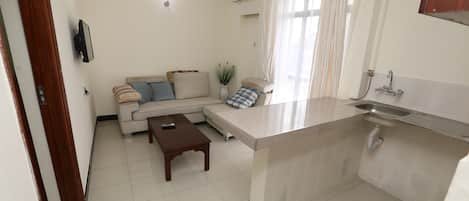 Um espaço de estar compacto com cozinha compacta, um sofá com almofadas, mesa de centro e TV fixada na parede.